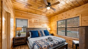 Quarto de cabana de madeira com cama, mesas de cabeceira, ventilador de teto e janela com persianas.
