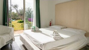 Quarto de hotel com uma cama grande, uma mesinha de cabeceira e vista para um jardim através de uma porta de vidro deslizante.