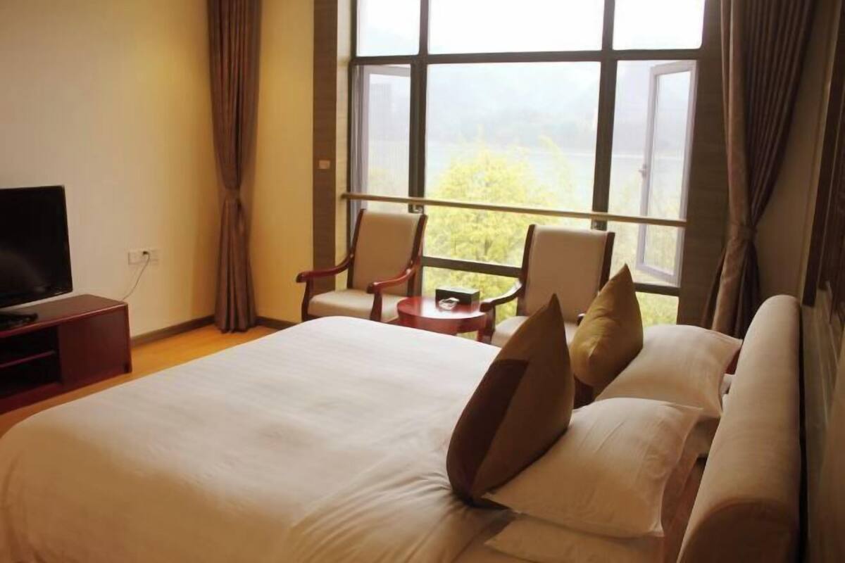 Quarto de hotel com uma cama grande, televisão, uma mesinha, duas cadeiras e janela com vista para o verde.