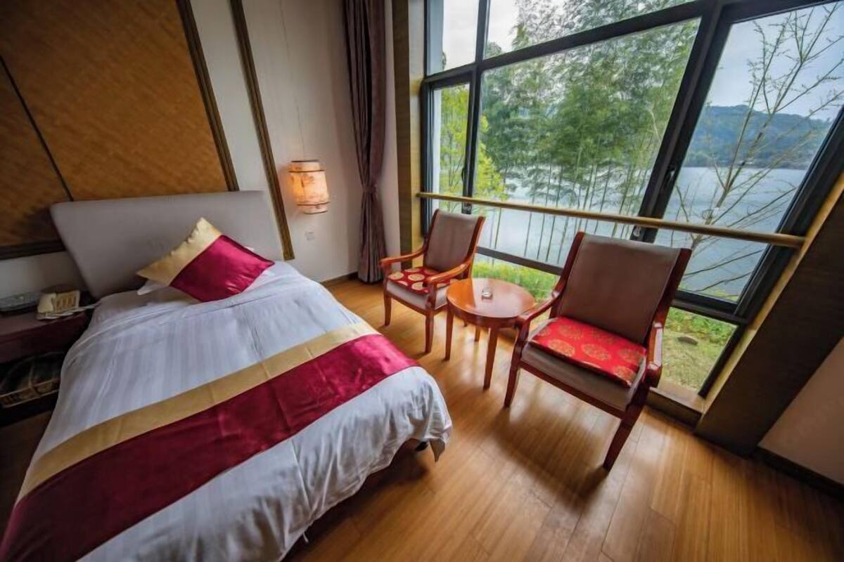 Quarto de hotel com uma cama grande, uma escrivaninha, duas cadeiras e vista para um lago através de grandes janelas.