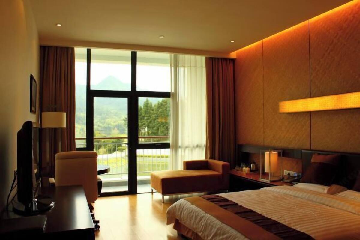 Quarto de hotel com cama, escrivaninha, cadeira e janela grande com vista para o verde.