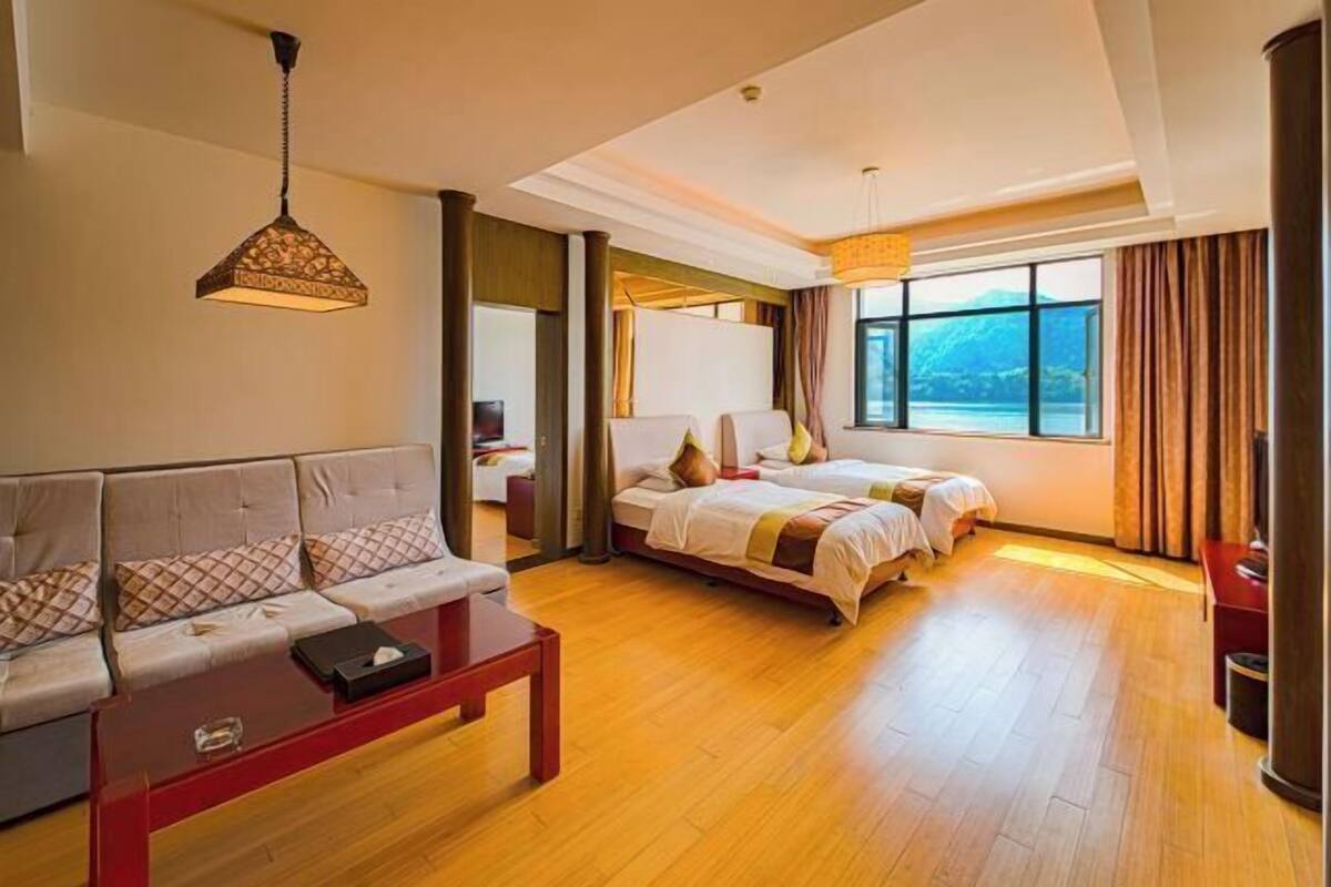 Quarto de hotel com sofá, duas camas, piso de madeira, uma janela ampla com vista e um lustre.