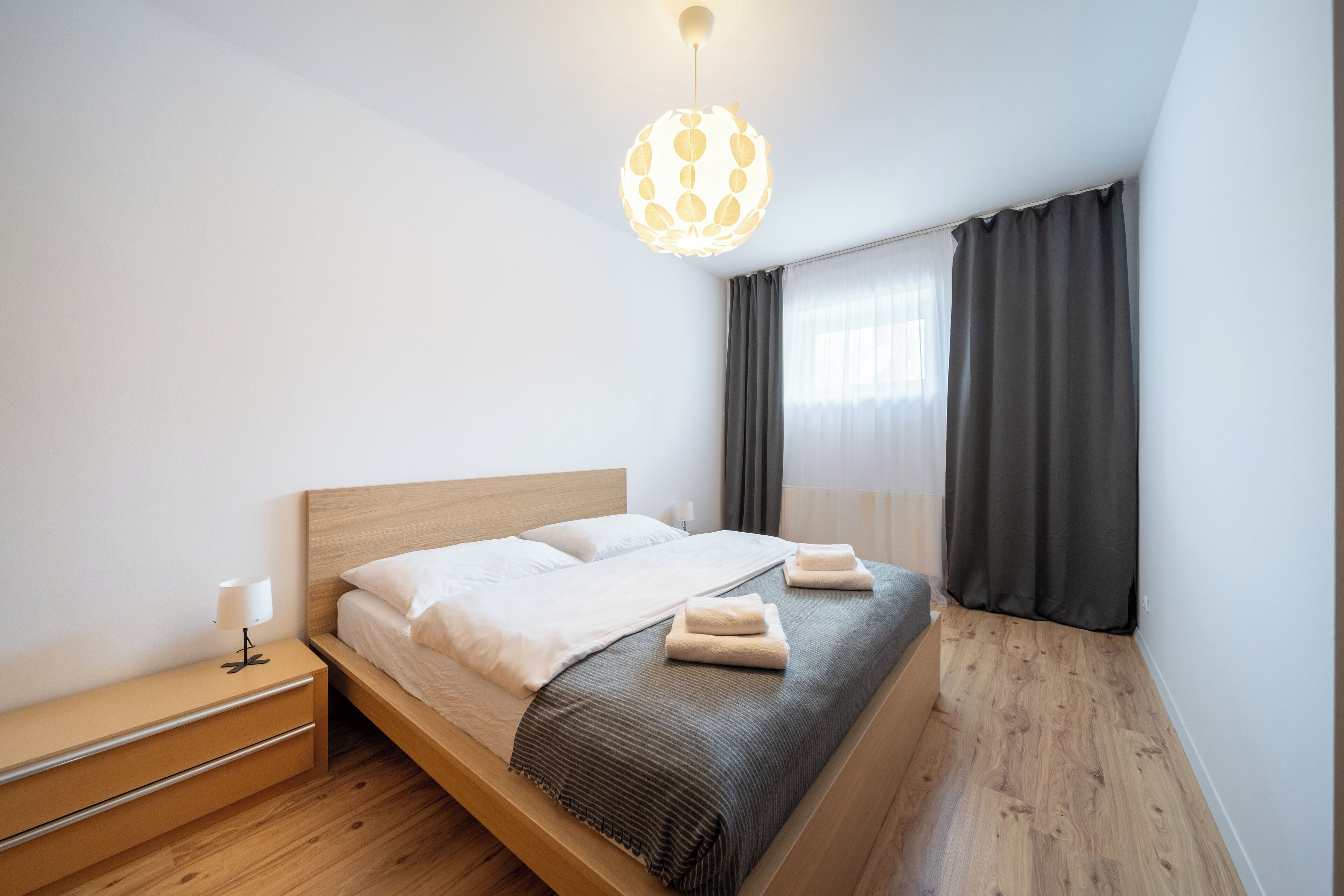 Um quarto moderno com cama de madeira, mesa de cabeceira com abajur, luminária pendente e vista para o exterior através de uma janela com cortinas.