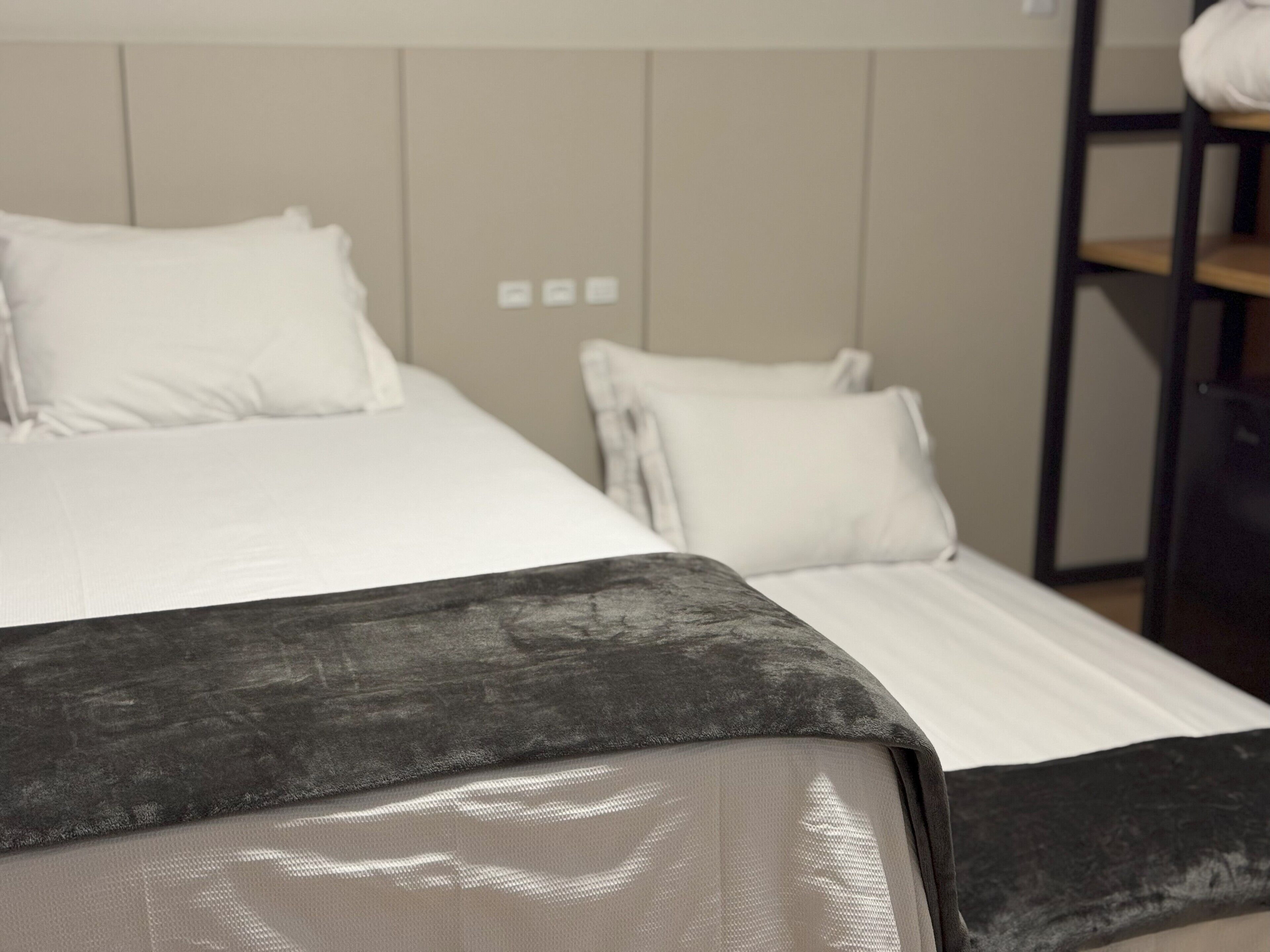 Quarto de hotel com duas camas, cada uma com roupa de cama e almofadas brancas. Possui cabeceira de madeira e uma prateleira com uma almofada branca.