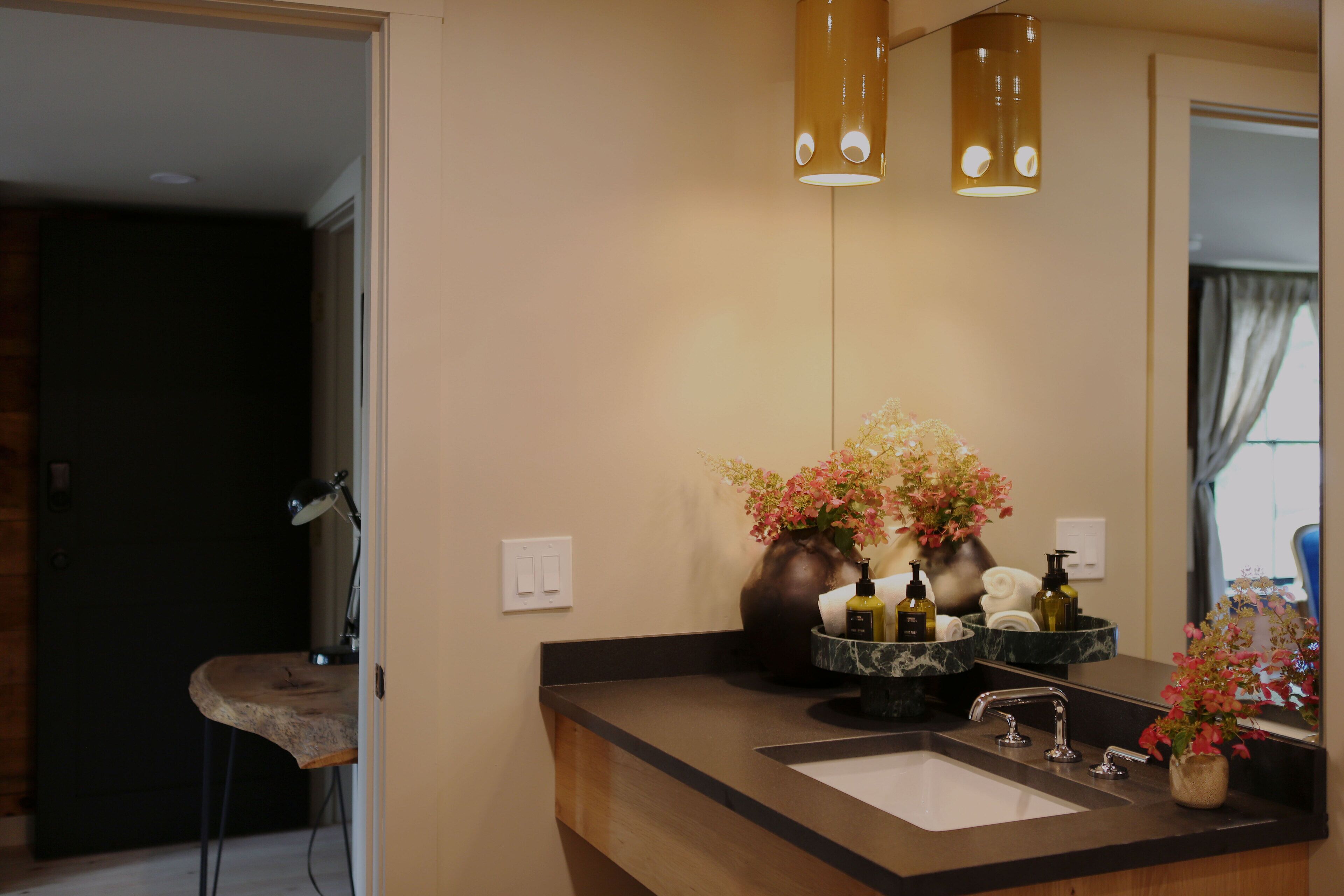 Um banheiro moderno com um espelho grande, uma pia e um lavatório. Há duas luminárias pendentes acima do espelho, e um vaso com flores no lavatório.