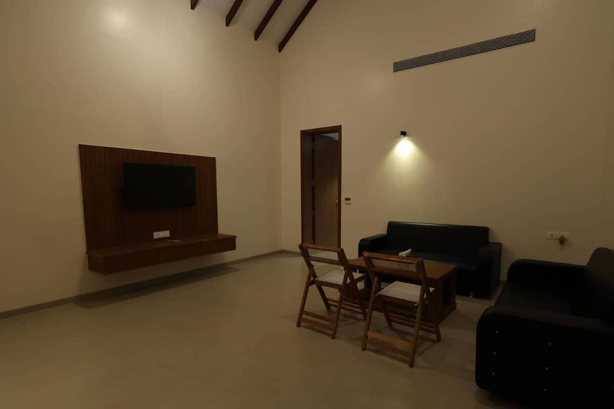 Uma sala de estar com televisão de tela plana na parede, uma mesa de jantar com cadeiras, um sofá preto e uma poltrona preta.