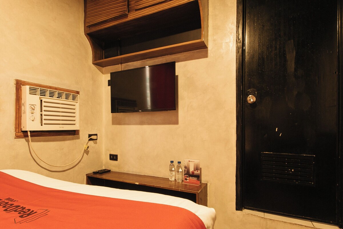 Quarto de hotel com cama, televisão fixada na parede, aquecedor, mesa de cabeceira com artigos de higiene pessoal e uma porta.