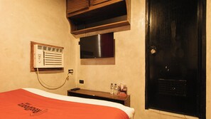 Quarto de hotel com cama, televisão fixada na parede, aquecedor, mesa de cabeceira com artigos de higiene pessoal e uma porta.