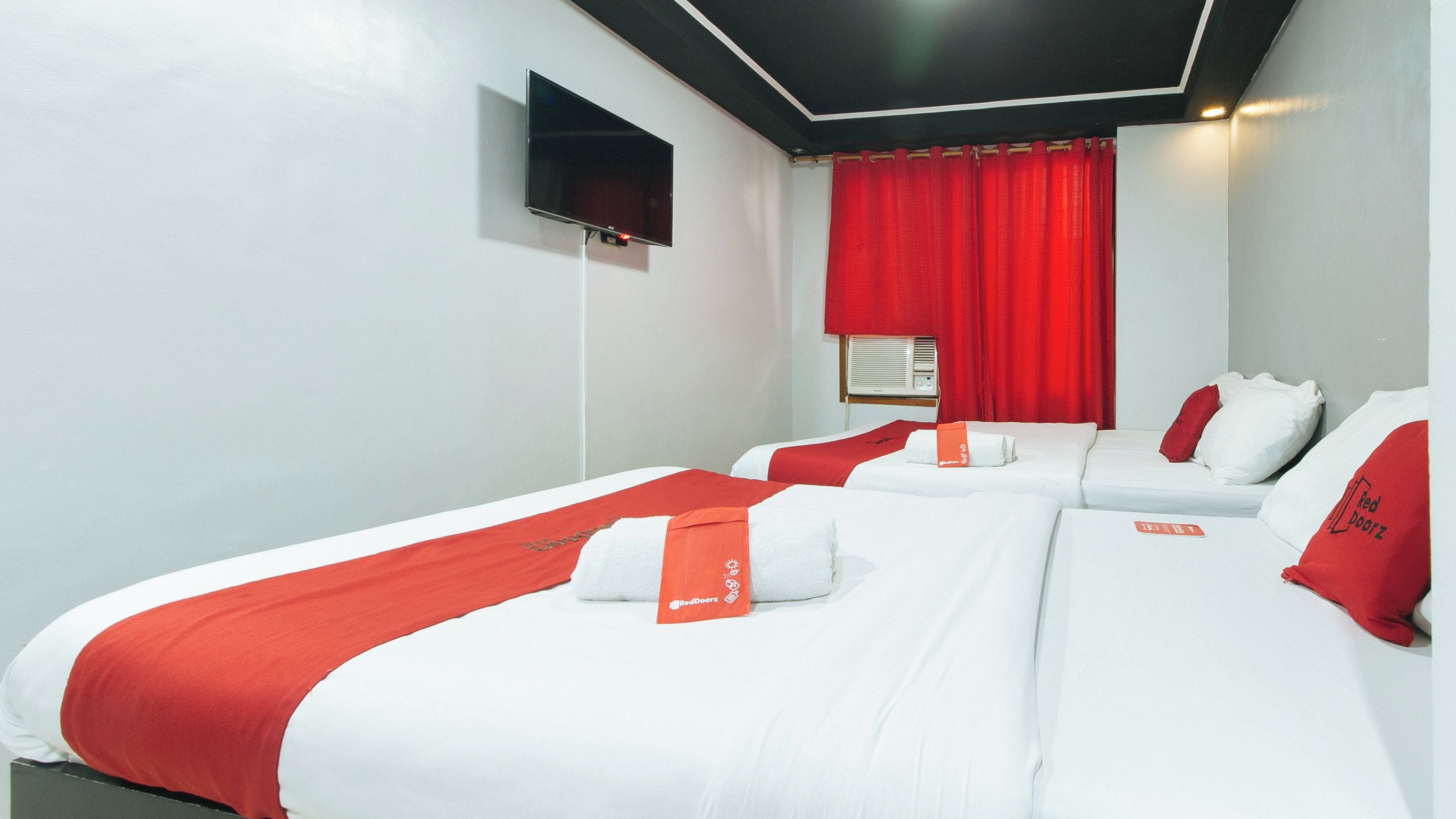 Quarto de hotel com duas camas, roupa de cama vermelha e branca, televisão de tela plana e cortina vermelha.