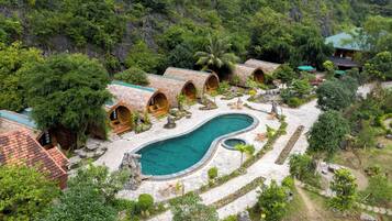 Resort com chalés de telhado de palha, piscina central e vegetação exuberante.