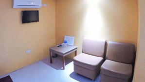 Um quarto pequeno com um sofá bege, uma mesinha com um controle remoto e um ar condicionado na parede.