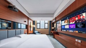 Quarto de hotel moderno com uma cama grande, uma mesa de trabalho, uma cadeira, uma TV na parede exibindo diversos canais e uma janela com vista para a cidade.