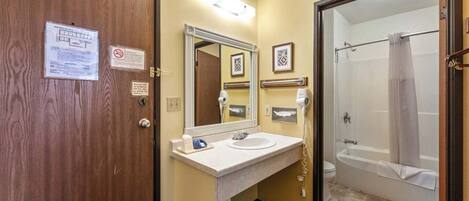 Casa de banho de hotel com porta de madeira, lavatório branco com espelho, chuveiro com porta de vidro e um porta-toalhas.
