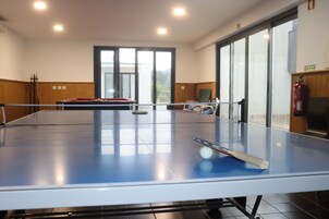 Uma mesa de tênis de mesa em um cômodo com piso de madeira, um extintor de incêndio e uma porta que dá para o exterior.