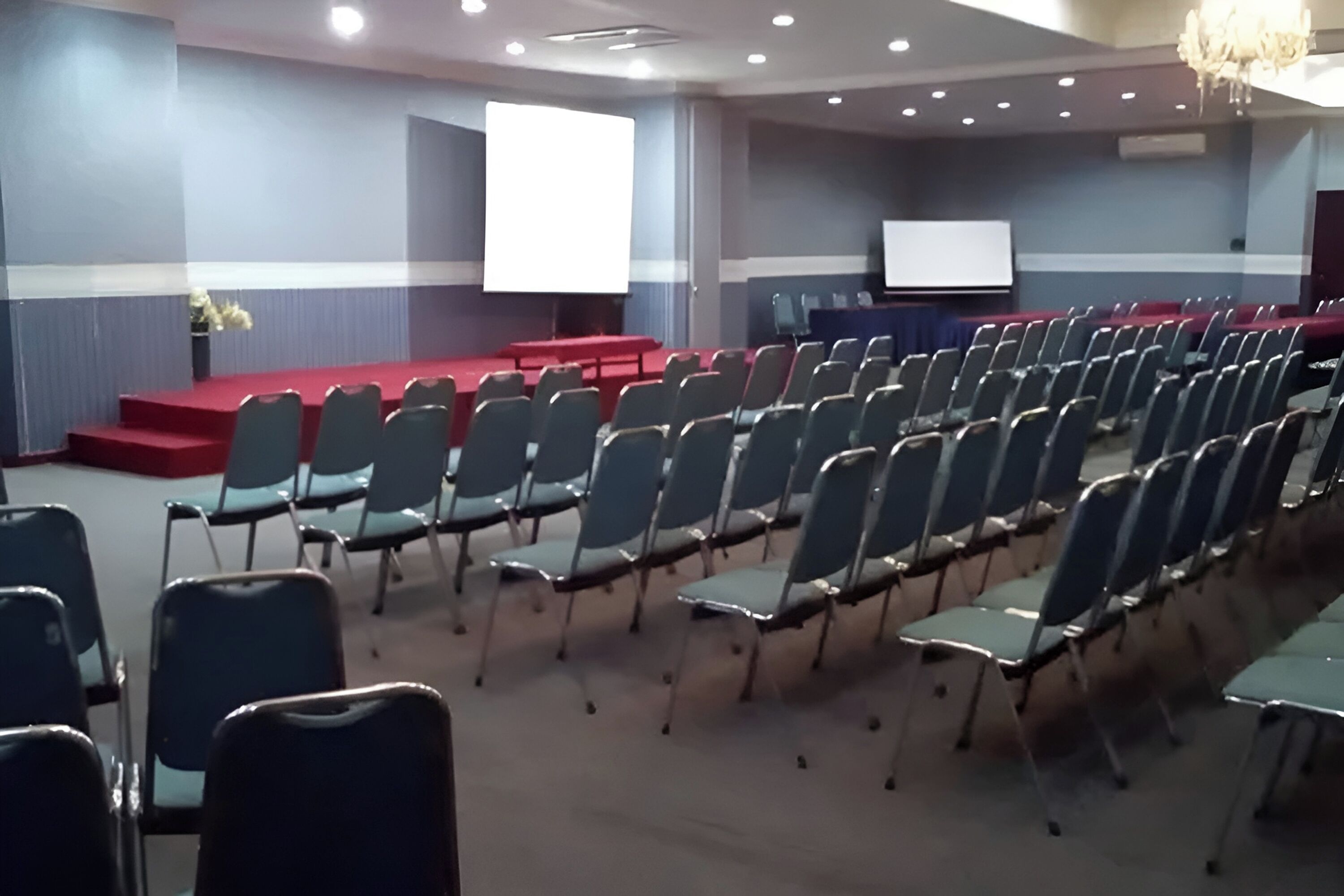 Uma sala de conferências com fileiras de cadeiras, um palco com tapete vermelho e uma tela de projetor.