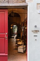Uma porta vermelha dá acesso a uma sala de jantar com mesas e cadeiras, um lustre e uma placa com a inscrição "LA CONCIA LOGANZA".