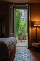 Quarto de hotel com cama, cadeira, mesa e porta aberta para área externa com vista para vegetação e um edifício.