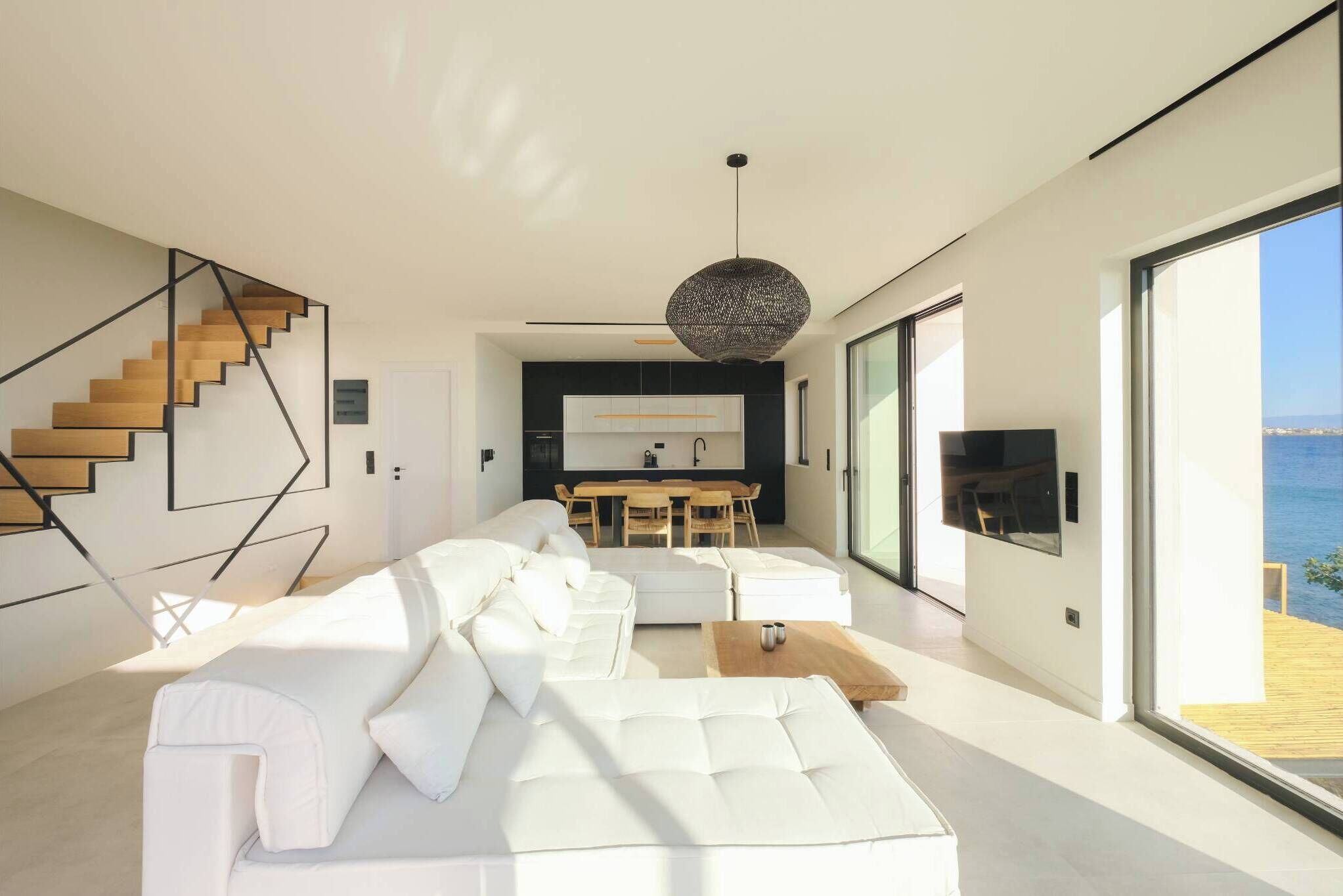 Uma sala de estar moderna com um sofá branco, uma área de jantar com mesa e cadeiras, uma cozinha com armários brancos, e uma grande janela com vista para o mar.
