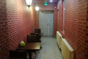 Um corredor estreito com paredes de tijolo vermelho, uma porta branca no final e mesas e cadeiras ao longo das laterais.