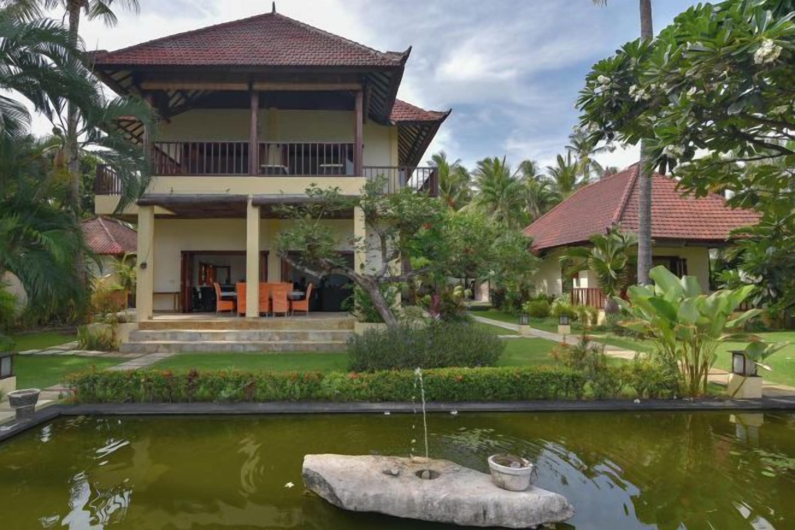 Um edifício de dois andares com varanda, rodeado por um jardim e um lago.