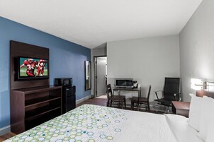 Quarto de hotel com cama, televisão, escrivaninha com cadeira, micro-ondas e frigorífico.