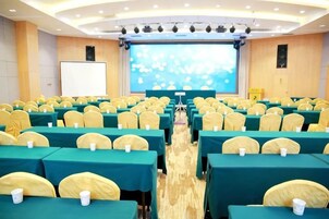 Sala de conferência com fileiras de mesas e cadeiras, uma tela grande na frente e um palco.