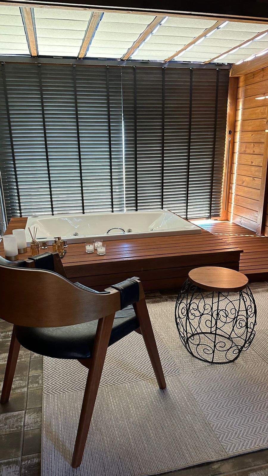 Um interior moderno com um banco de madeira, uma mesa redonda de metal e uma janela grande com persianas.