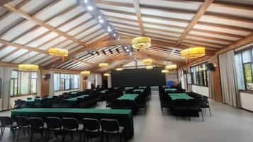 Sala de conferências com mesas, cadeiras, palco e teto alto com vigas de madeira e lustres.
