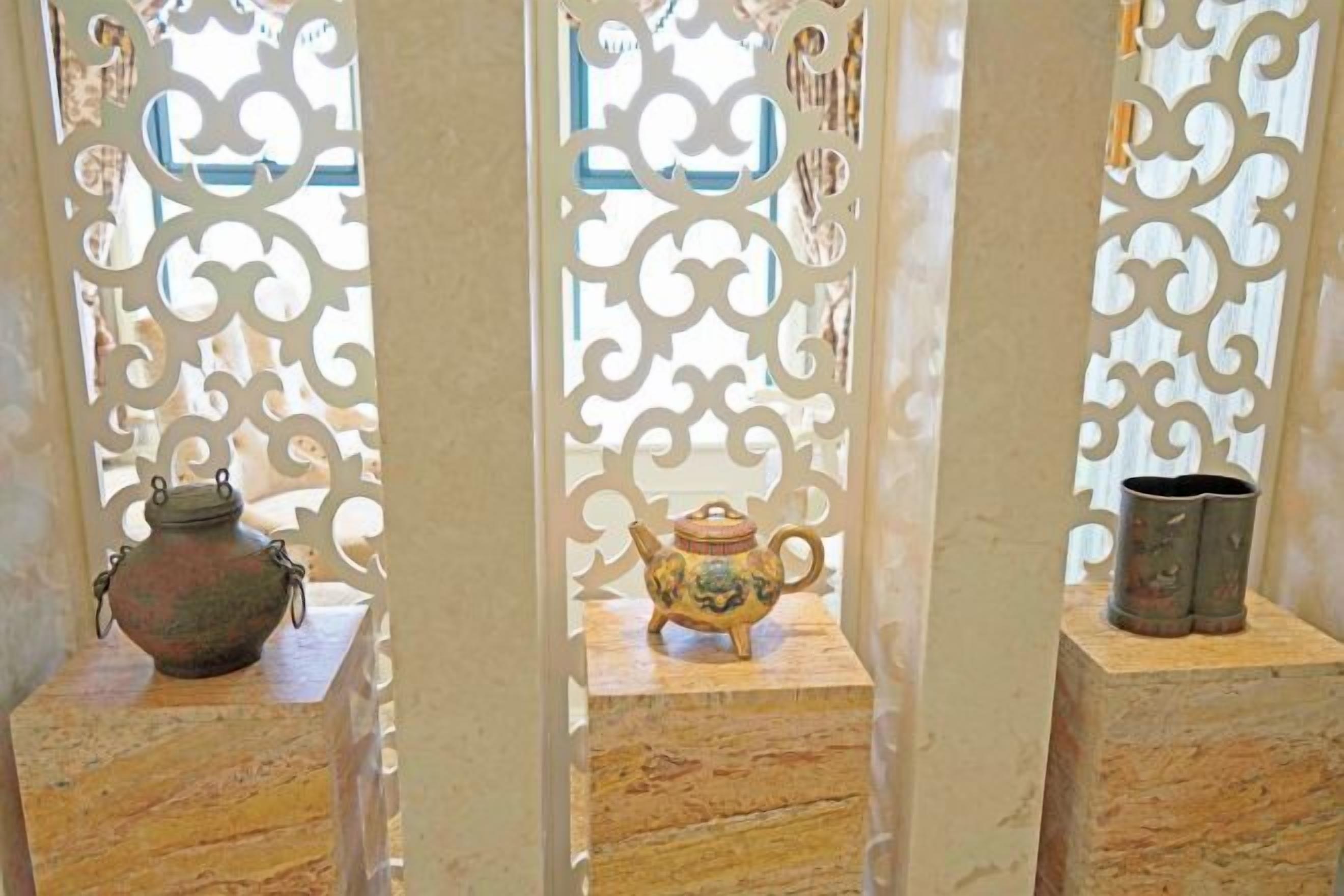 Quarto com um painel decorativo com padrões elaborados. Há duas mesinhas, cada uma com um objeto decorativo: um vaso de cerâmica e um pequeno vaso.