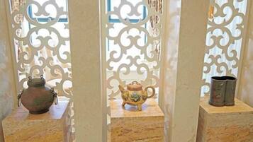 Quarto com um painel decorativo com padrões elaborados. Há duas mesinhas, cada uma com um objeto decorativo: um vaso de cerâmica e um pequeno vaso.