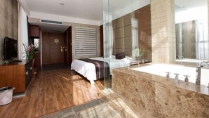 Quarto de hotel moderno com uma cama grande, divisória de vidro, banheira de mármore e uma televisão sobre um armário de madeira.