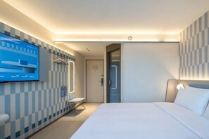 Quarto de hotel moderno com uma cama, televisão fixada na parede, uma porta e uma mesinha.