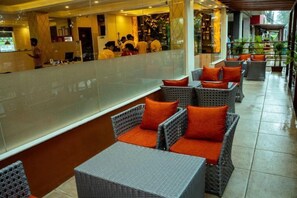 Um hall de hotel com móveis de vime e almofadas laranja, uma área de recepção com funcionários e um painel de vidro.