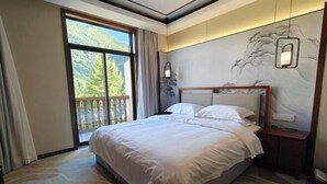 Quarto de hotel com uma cama grande, mesa de cabeceira, luminária na parede e vista para o exterior pela janela.