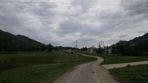 Uma estrada rural serpenteando por uma paisagem verde com montanhas ao longe e alguns edifícios.