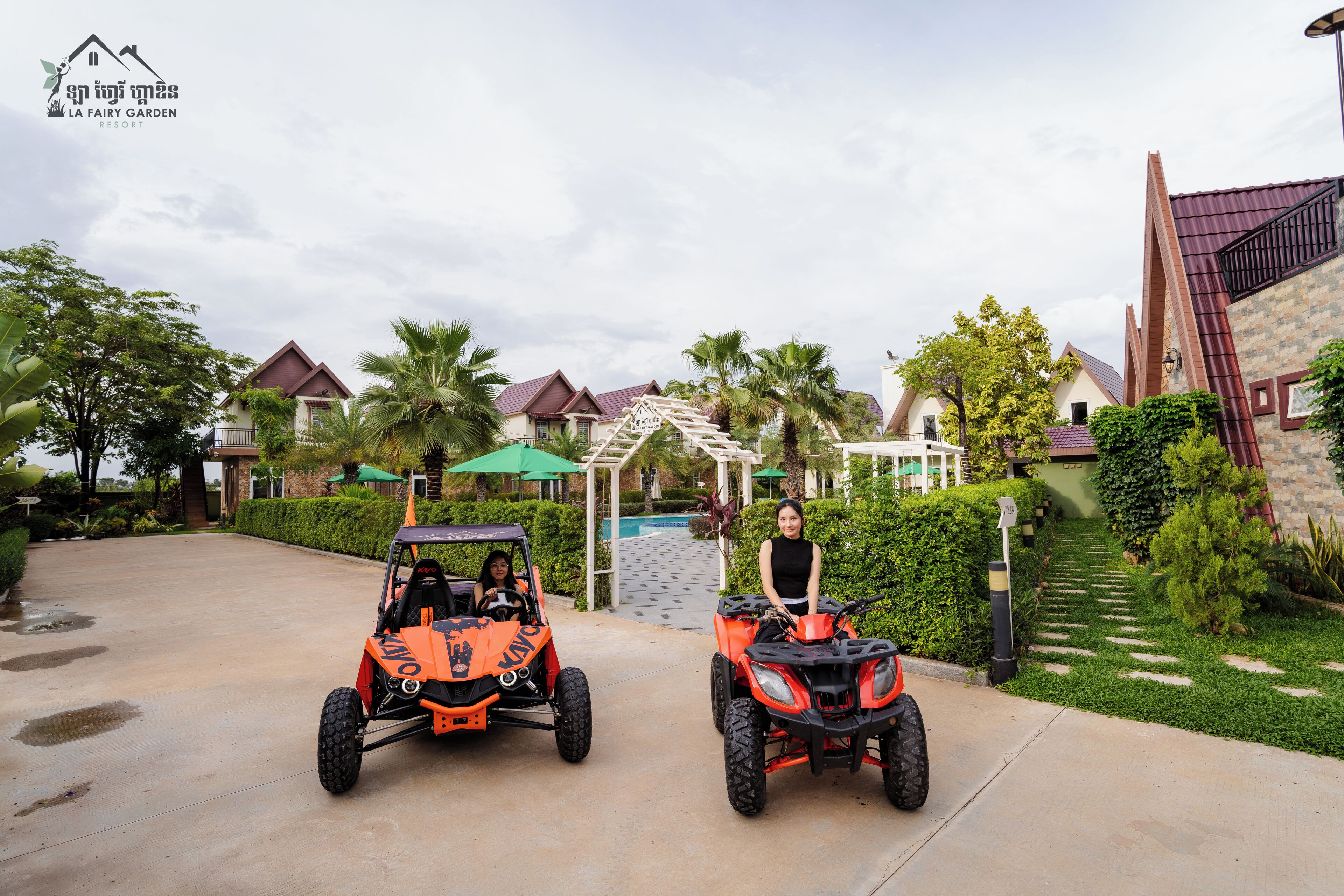 Duas motos de aventura laranja estacionadas em um caminho pavimentado em área de resort, com palmeiras e piscina ao fundo.