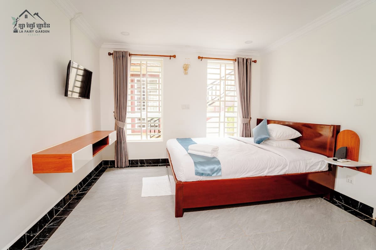 Quarto de hotel com cama, televisão na parede, mesa de cabeceira de madeira e janela com cortinas.