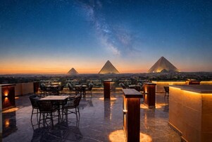Terraço no telhado com mesas, cadeiras, área de bar e as pirâmides ao fundo.