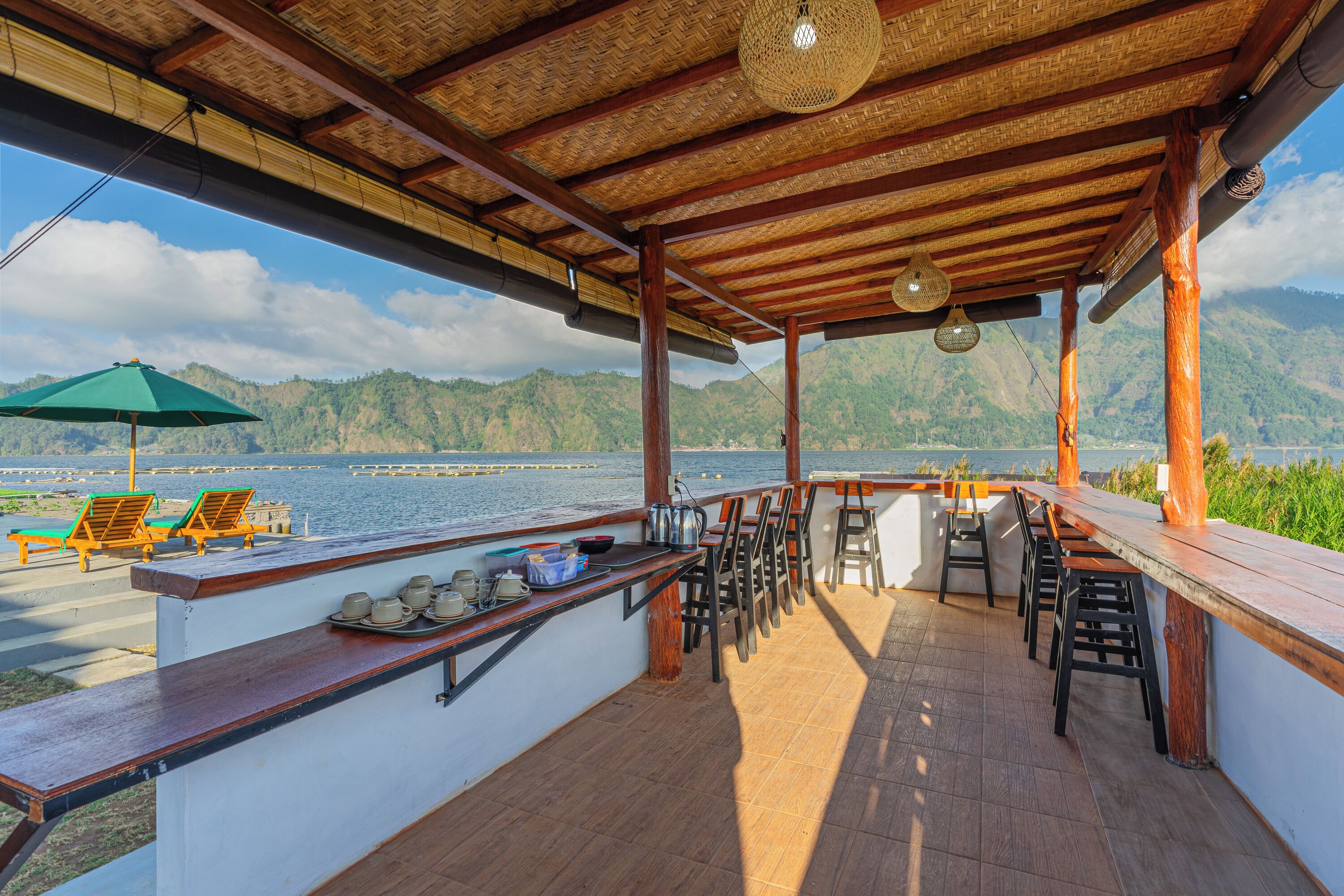 Uma área de bar ao ar livre coberta, com mobiliário de madeira, vista para o mar e montanhas ao fundo.