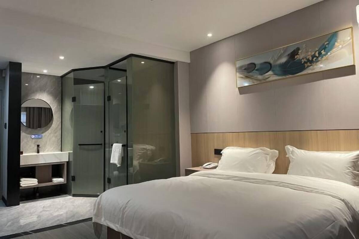 Quarto de hotel moderno com cama, chuveiro em vidro, toucador com espelho e um quadro na parede.
