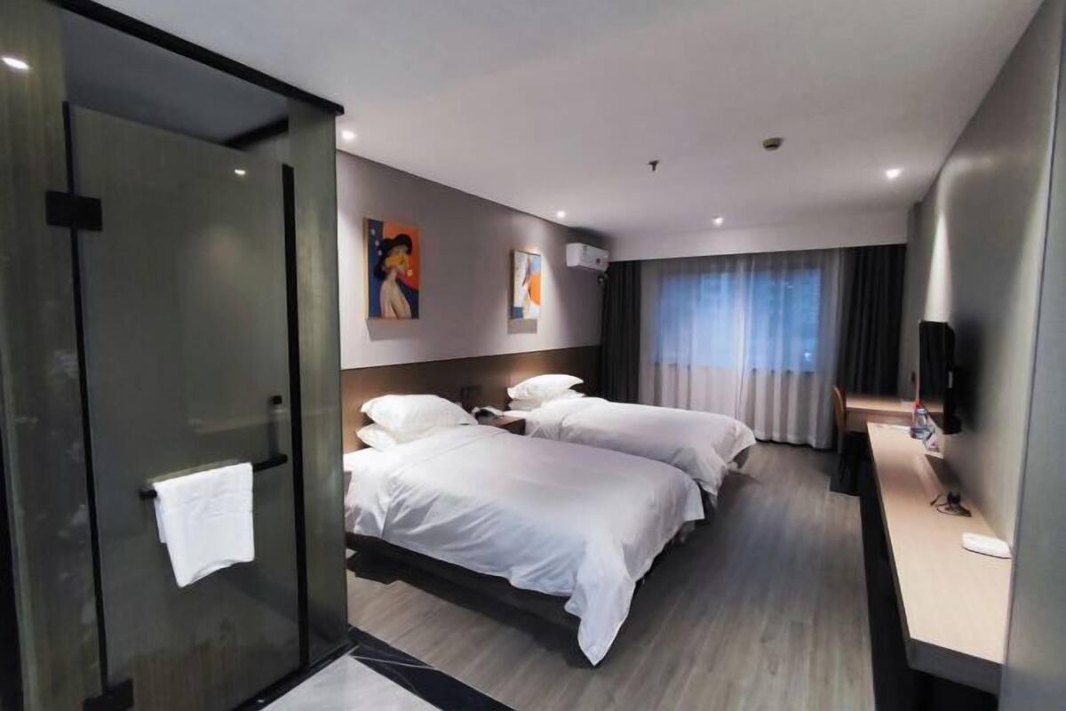 Quarto de hotel com duas camas, uma escrivaninha, uma cadeira, uma televisão e box de chuveiro de vidro.