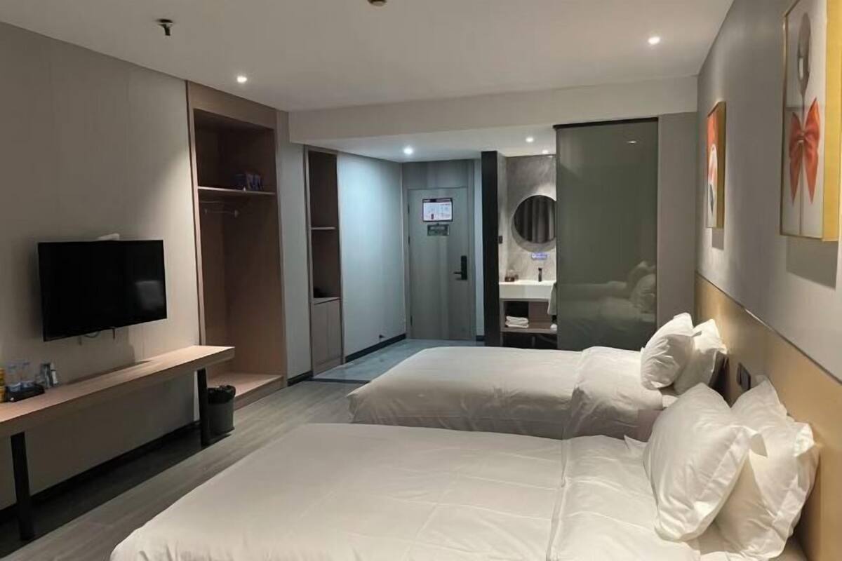 Quarto de hotel com duas camas, televisão, guarda-roupa e banheiro visível através de uma porta de vidro.