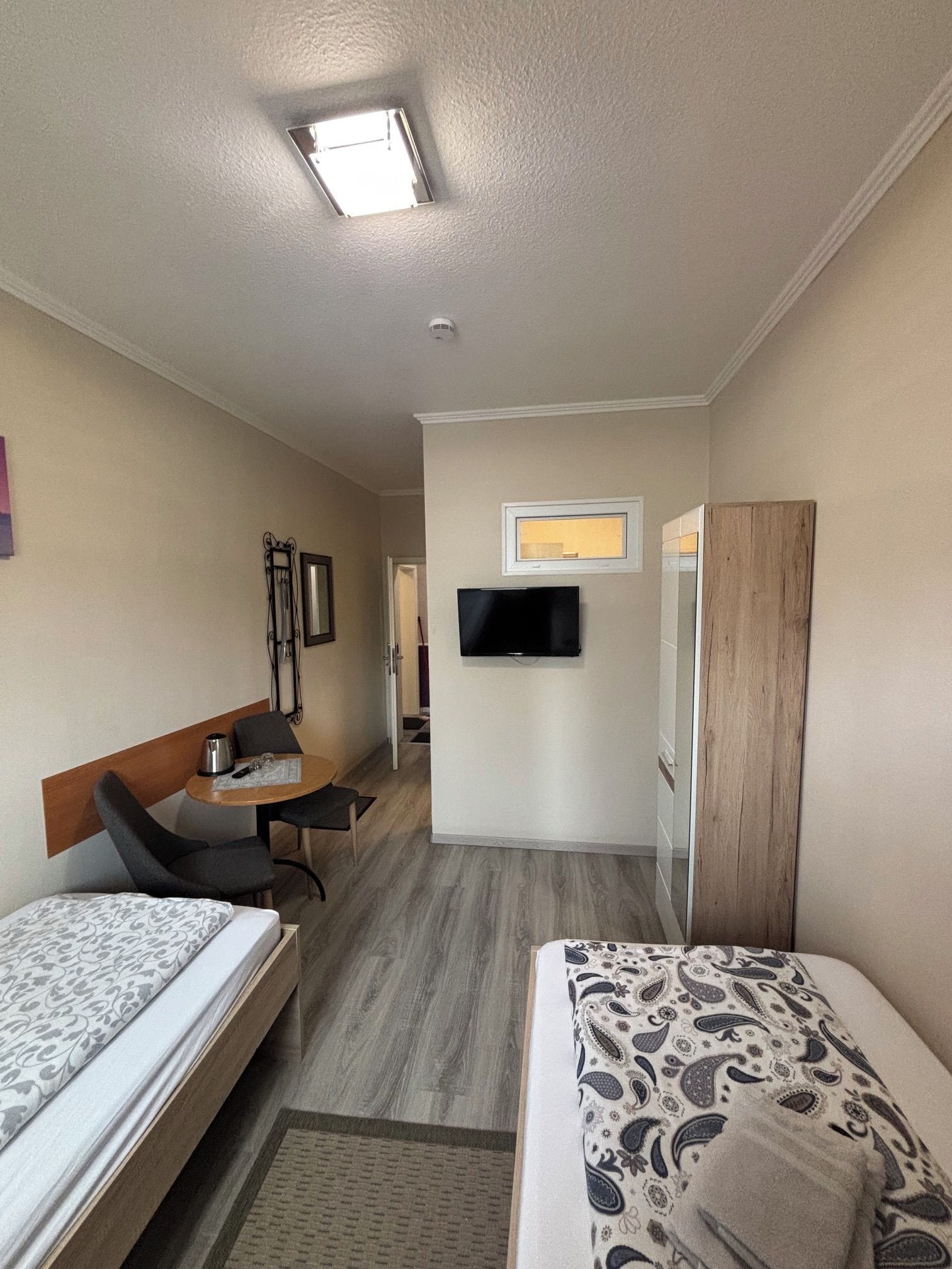Quarto de hotel com duas camas, uma escrivaninha com cadeira, televisão na parede e guarda-roupa de madeira.