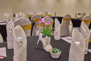 Uma sala de conferências com mesas redondas, cadeiras brancas com fitas amarelas e pequenos vasos com flores.