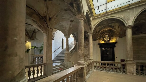 Interior com colunas de pedra, piso xadrez e escadaria que leva a outro nível.