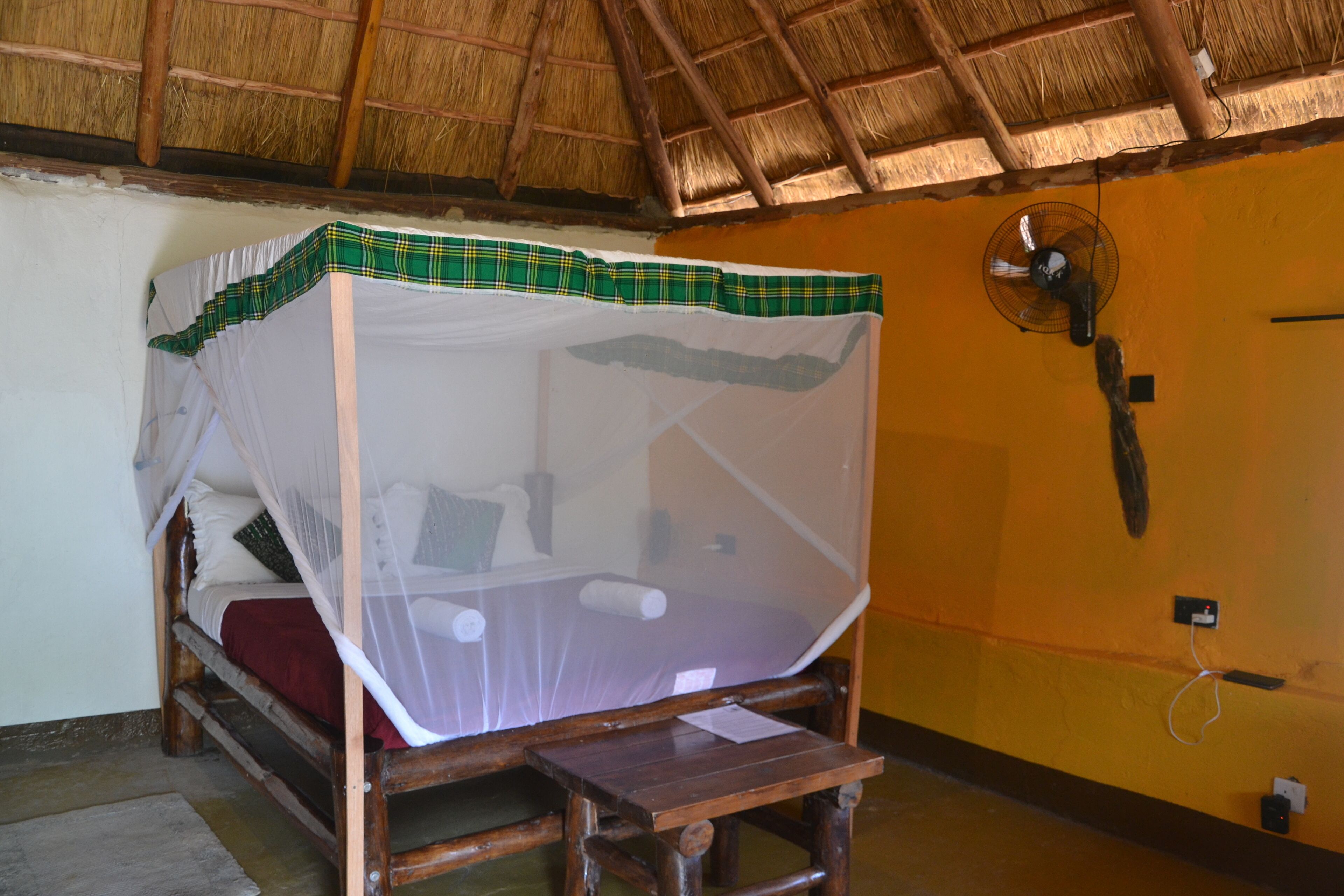 Uma cama de solteiro com mosquiteiro, móveis de madeira, um ventilador e uma tomada elétrica numa parede amarela.