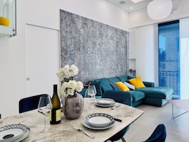 Uma sala de estar moderna com um sofá azul-turquesa, uma grande parede com arte abstrata e uma área de jantar com mesa de mármore posta para refeição.