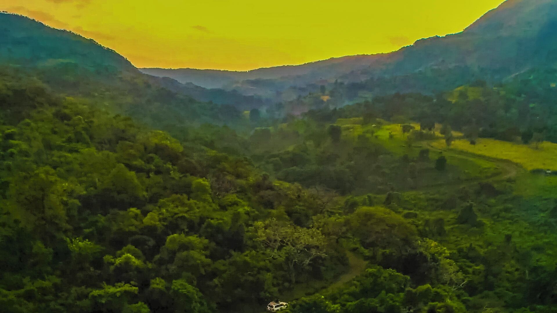 Um vale verdejante com uma estrada sinuosa e um veículo em primeiro plano.