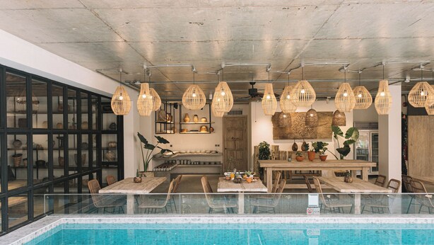 Área de piscina com barreira de vidro, luminárias pendentes de vime e espaço de estar com mesas e cadeiras.
