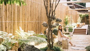 Mulher sentada em um banco de pedra em um jardim com cerca de bambu, caminhos de pedra e diversas plantas.